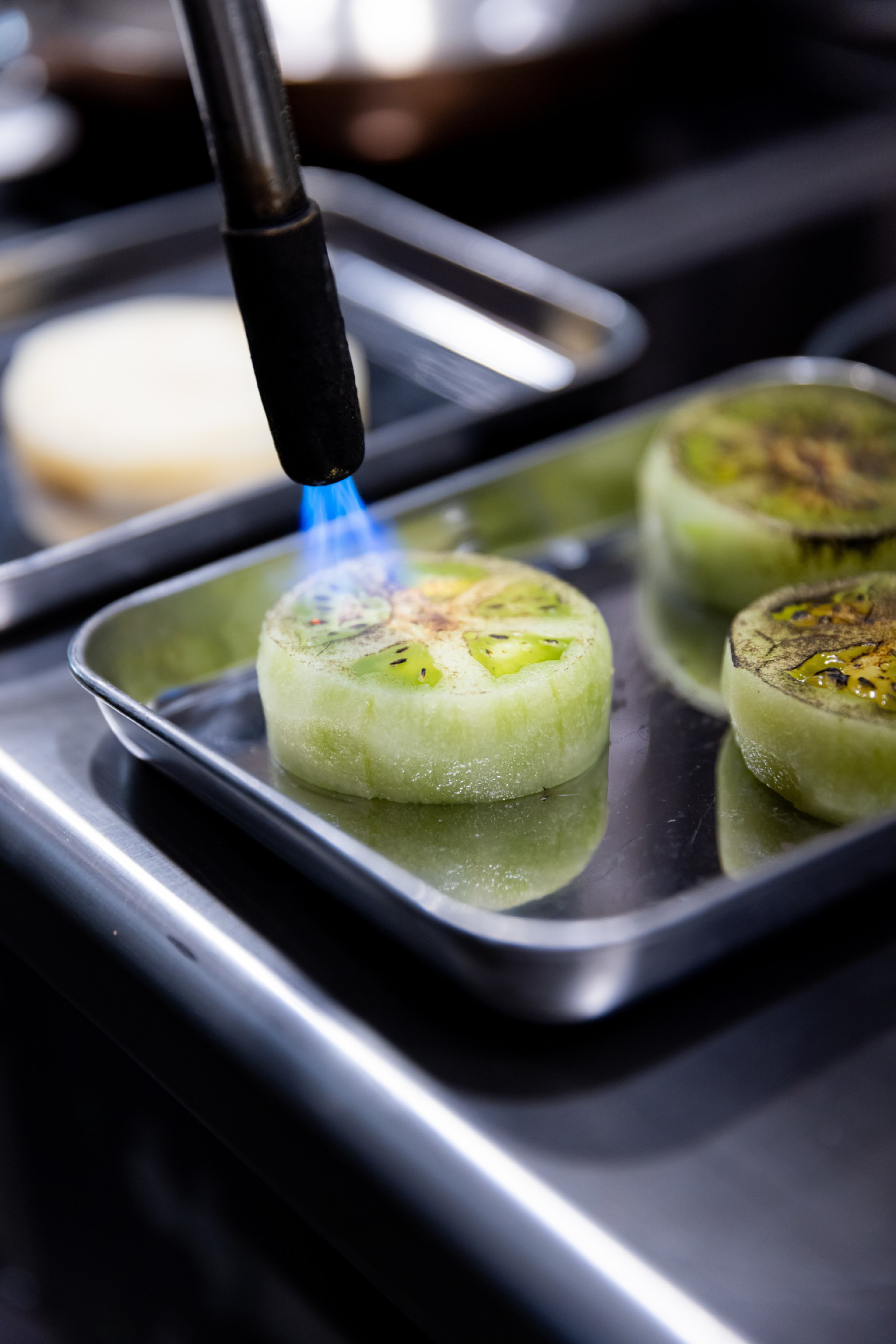 A kitchen torch is caramelizing the top of a green tomato slice on a ...