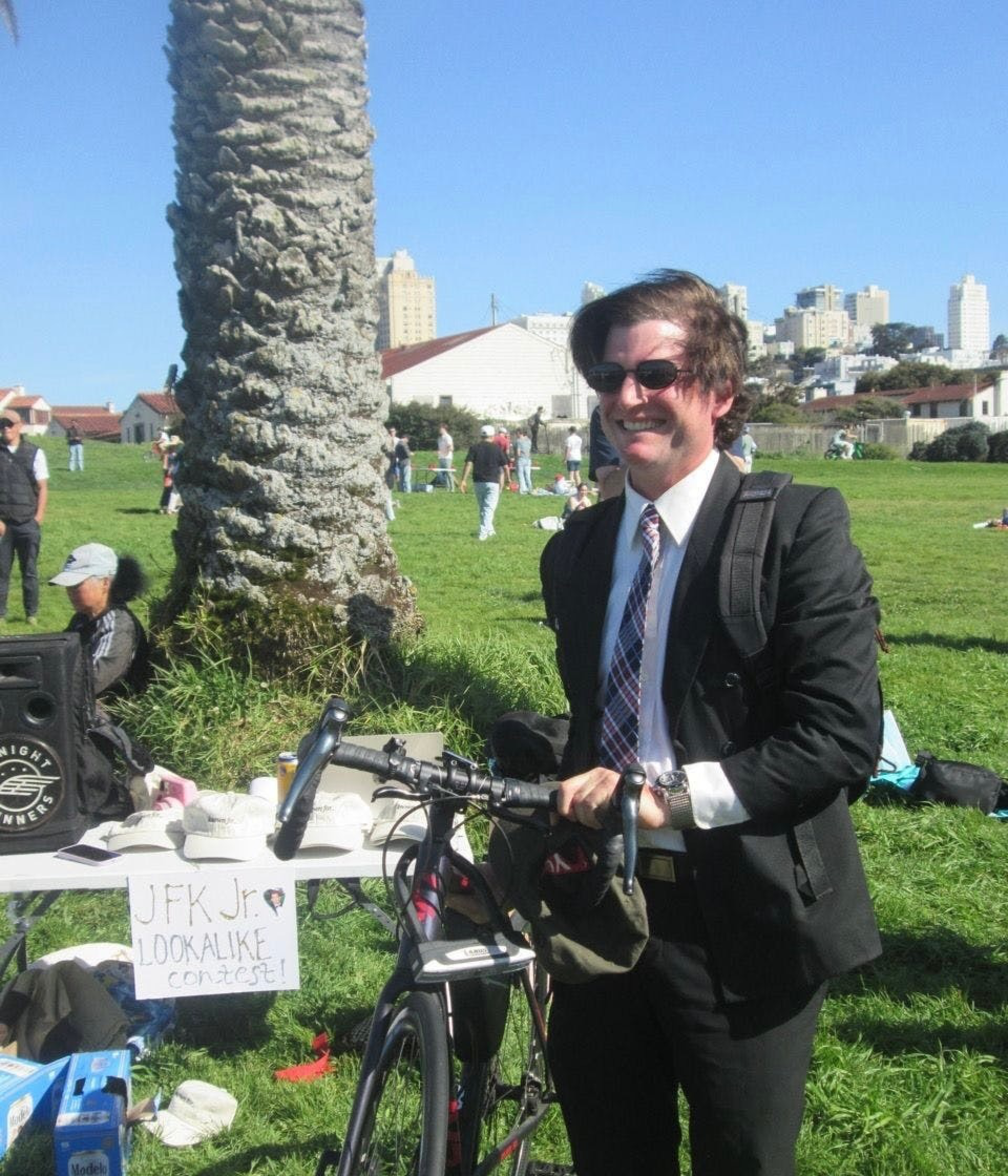 A man in a suit, tie, and sunglasses smiles while holding a bicycle at ...
