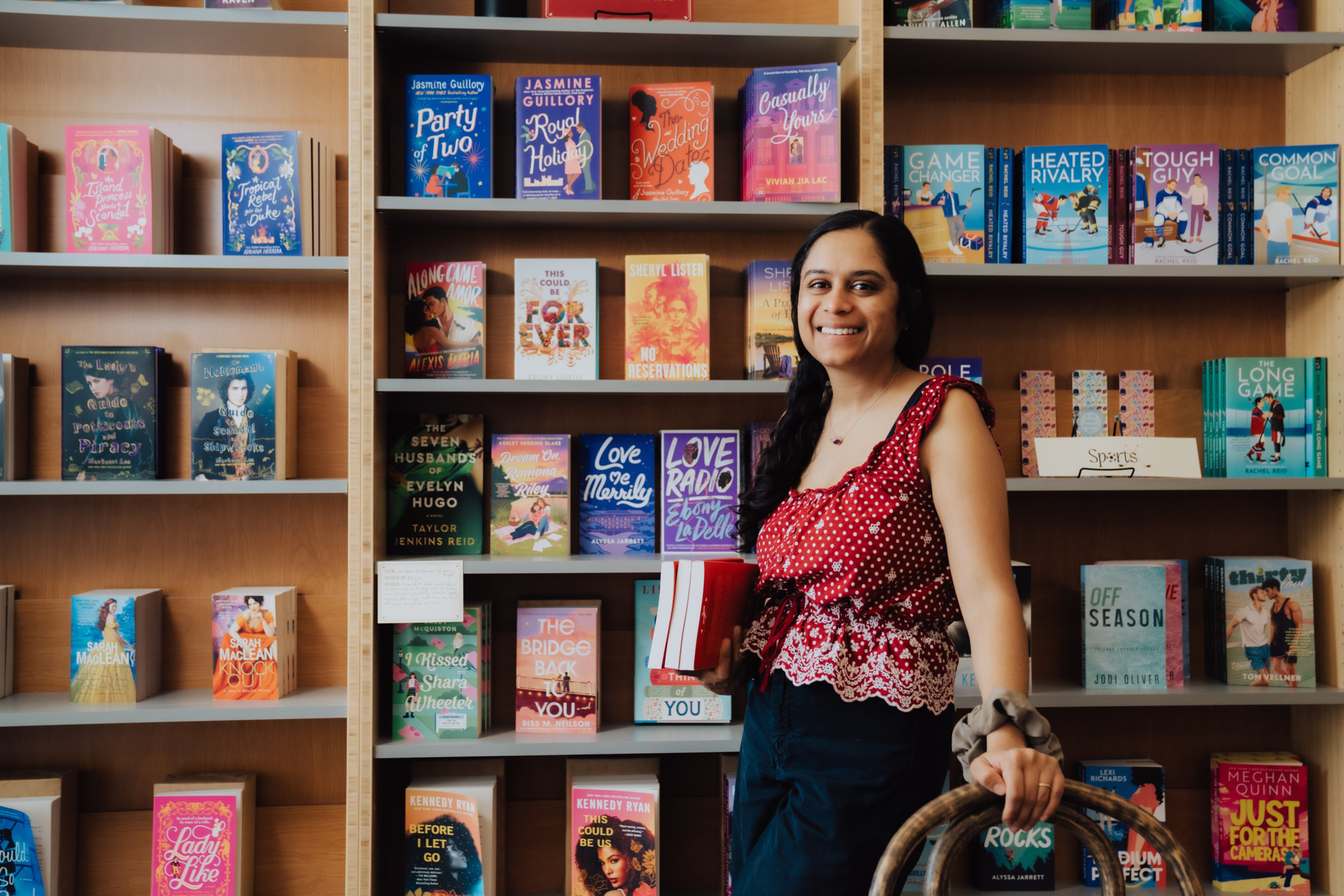 A smiling woman in a red polka-dot top holds books standing in front of ...