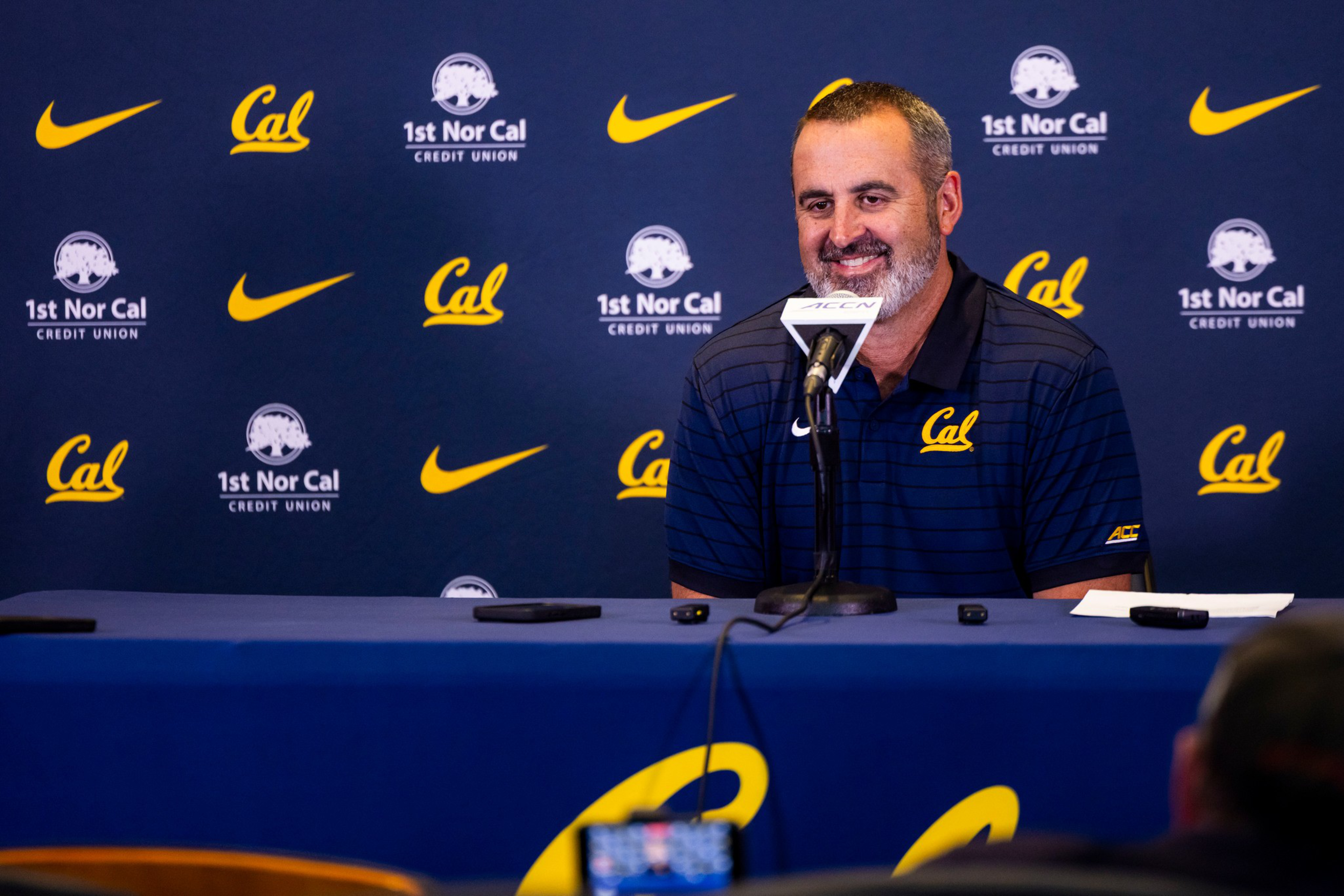 A man in a Cal polo shirt smiles while speaking into a microphone at a ...
