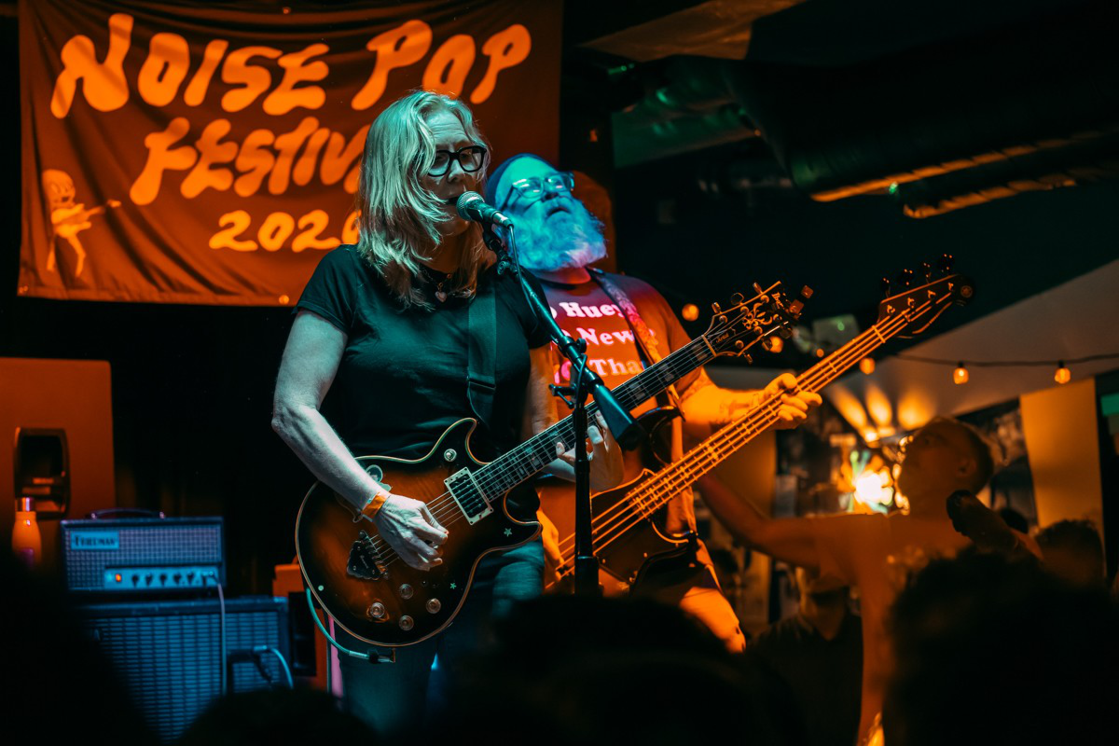 A female guitarist and a bearded bassist perform on stage under a ...