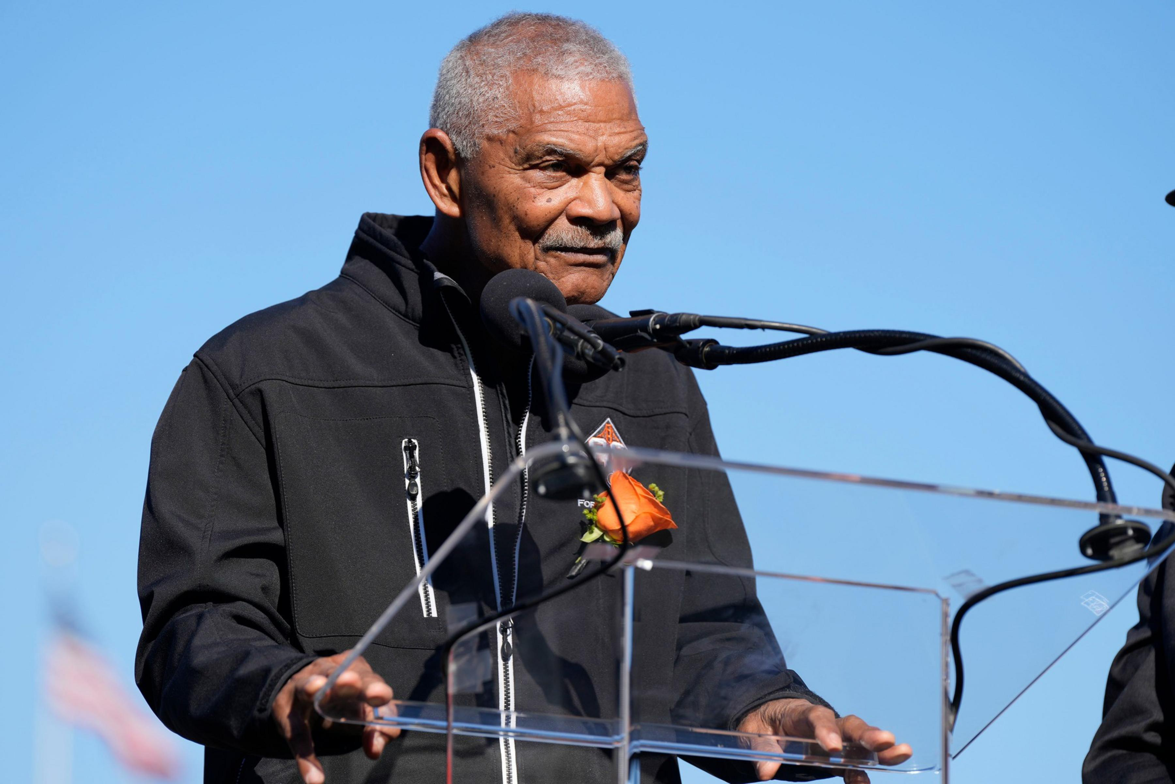 An older man with gray hair and a mustache speaks into microphones ...