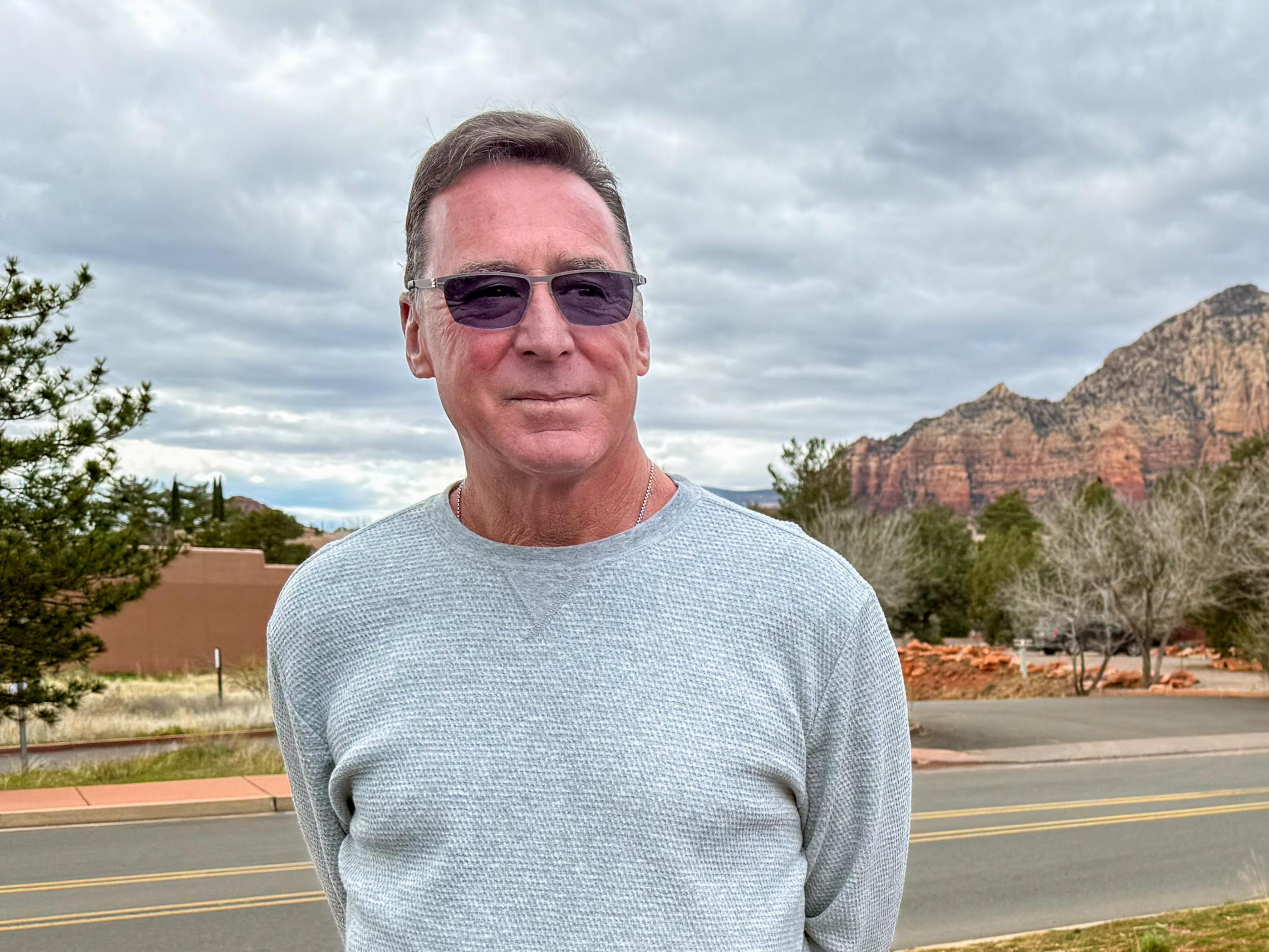 A man wearing a light gray sweater and dark sunglasses stands near a ...