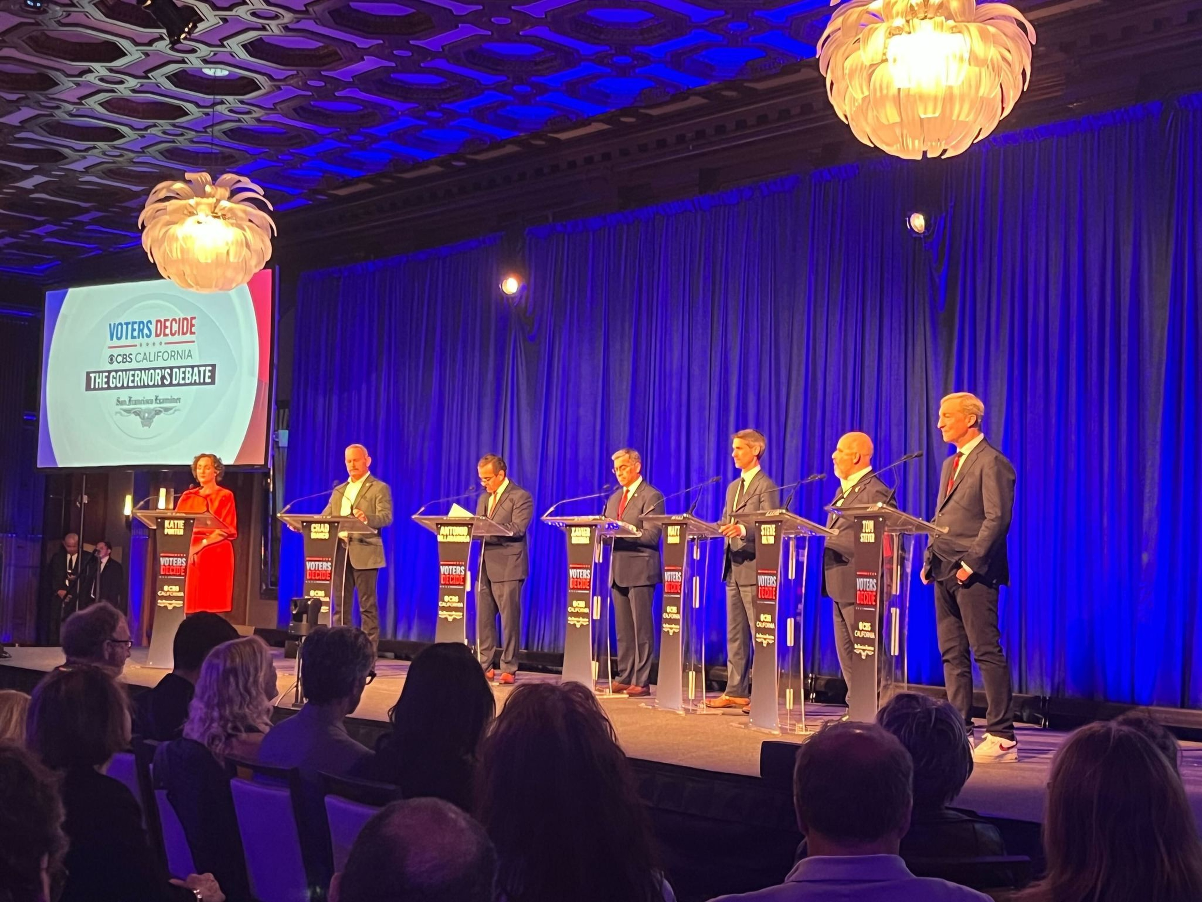 Seven candidates stand at individual podiums on a stage with blue ...