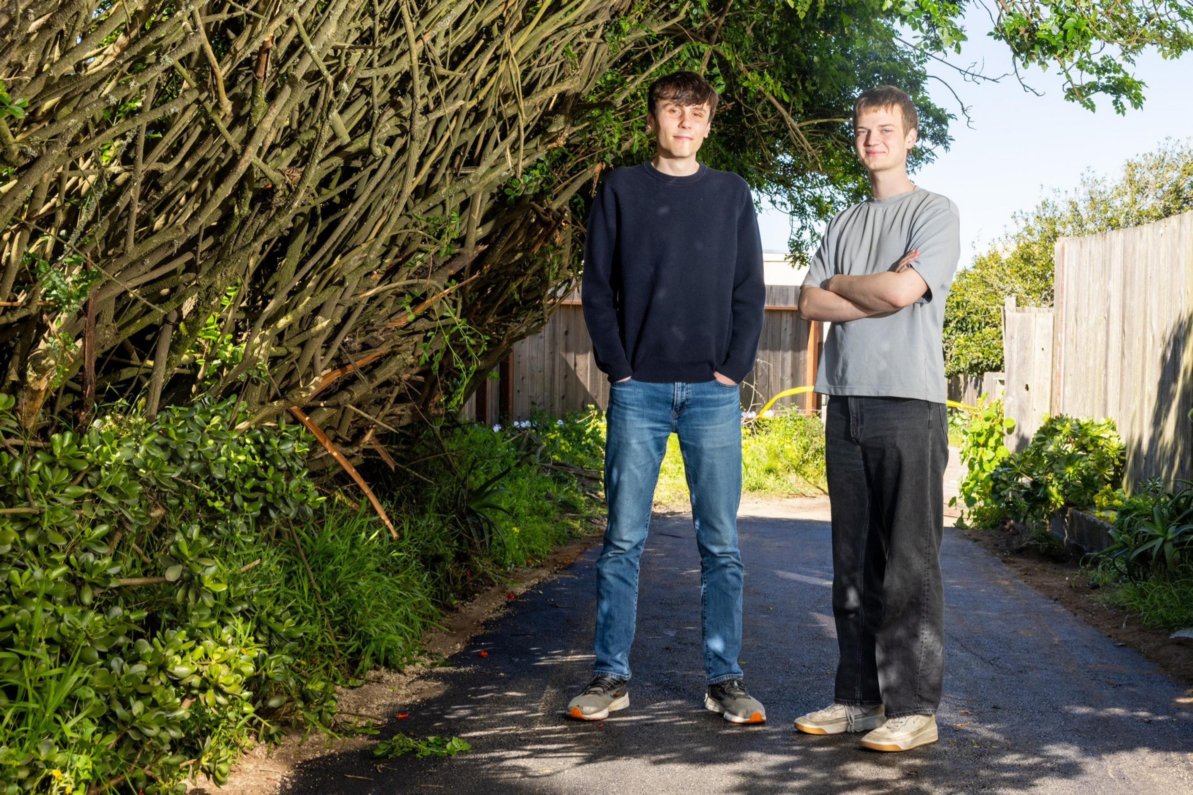 Two young men stand on a paved path between dense green bushes and a ...