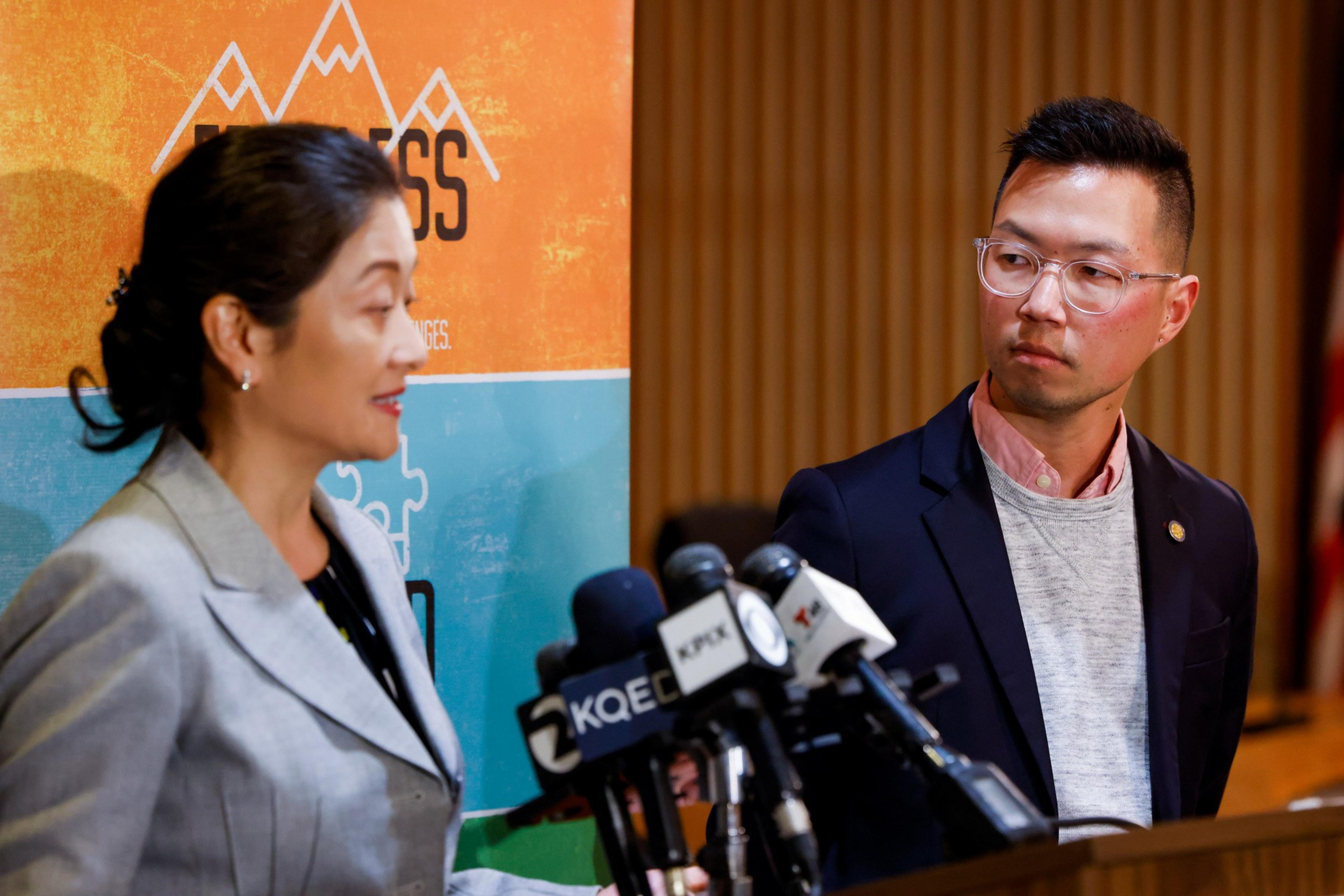 Two people stand at a podium with microphones. The woman speaks, while ...