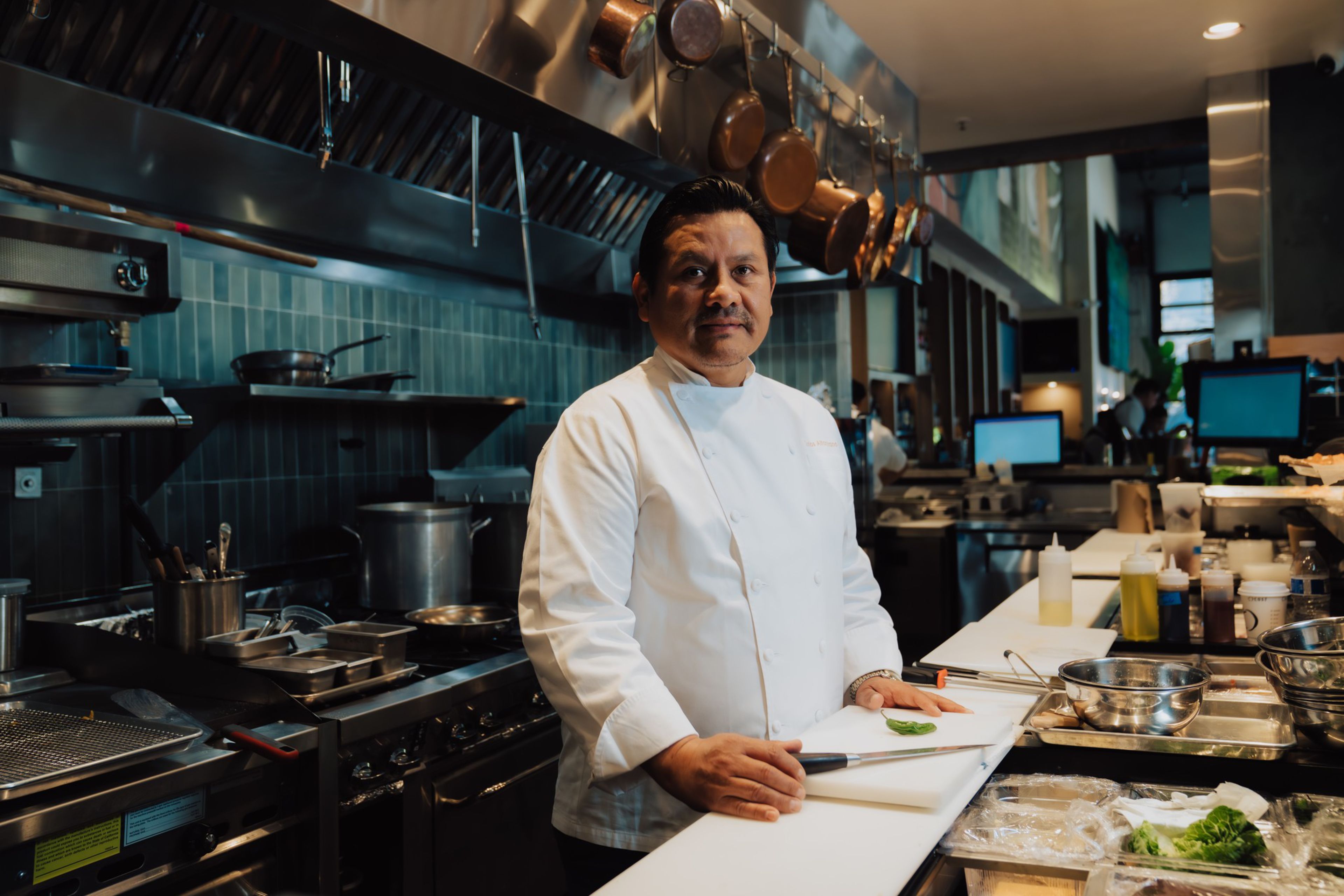 A chef in a white coat stands in a commercial kitchen, holding a knife ...
