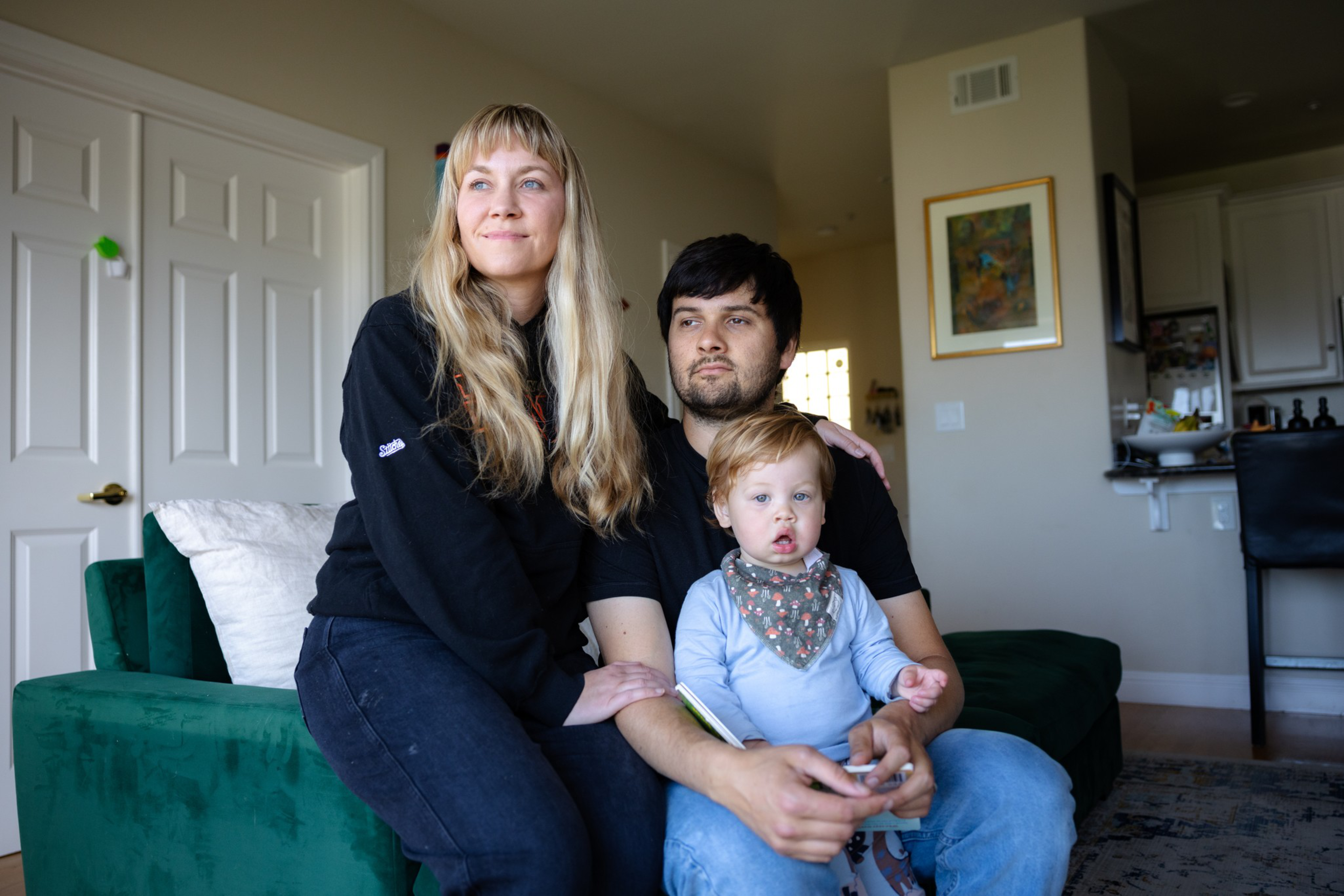 A woman, man, and toddler sit closely on a green couch in a cozy living ...