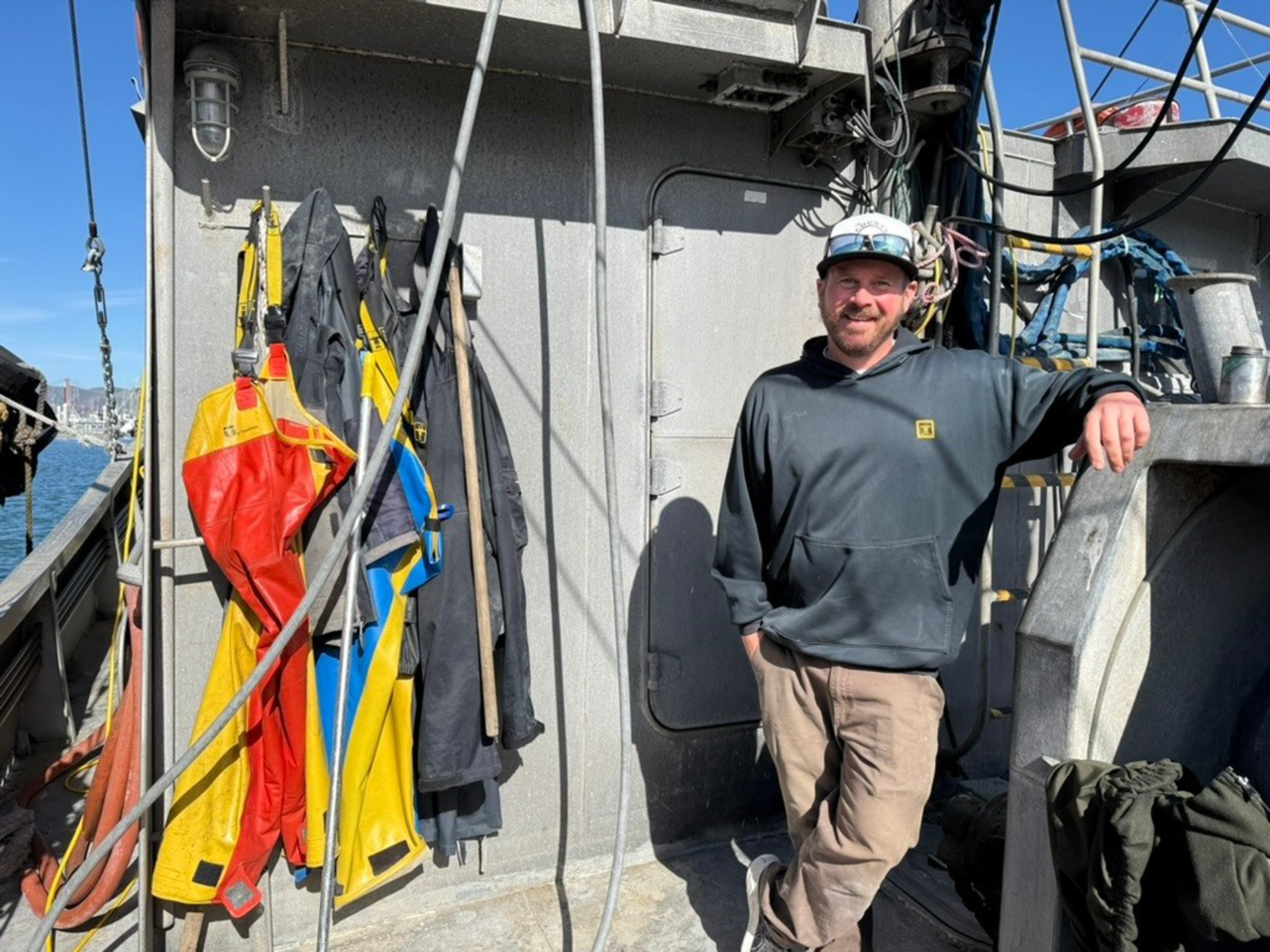 A man in a black hoodie and cap leans against a gray wall on a boat ...