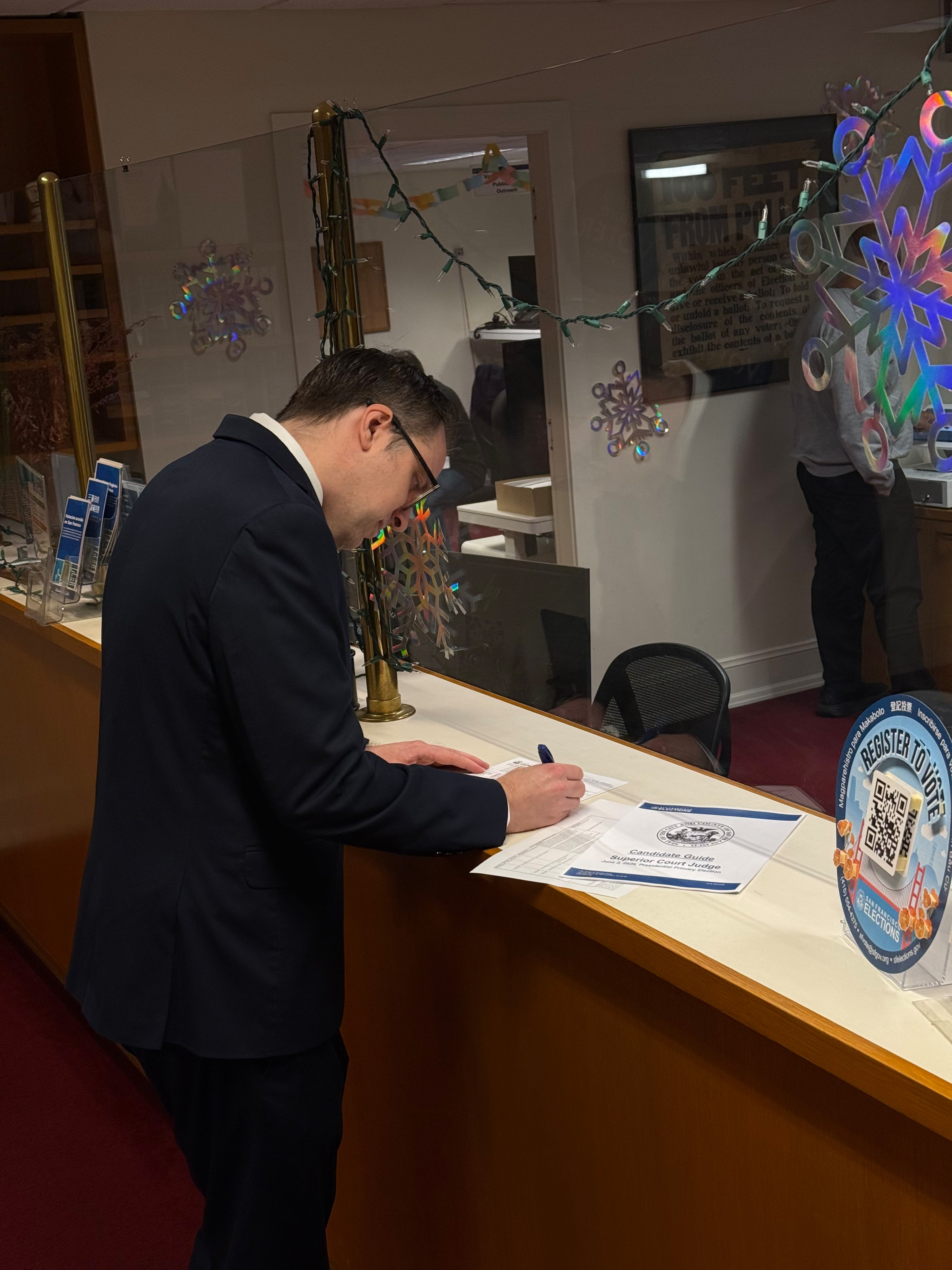 A man in a dark suit and glasses is signing a document at a counter ...