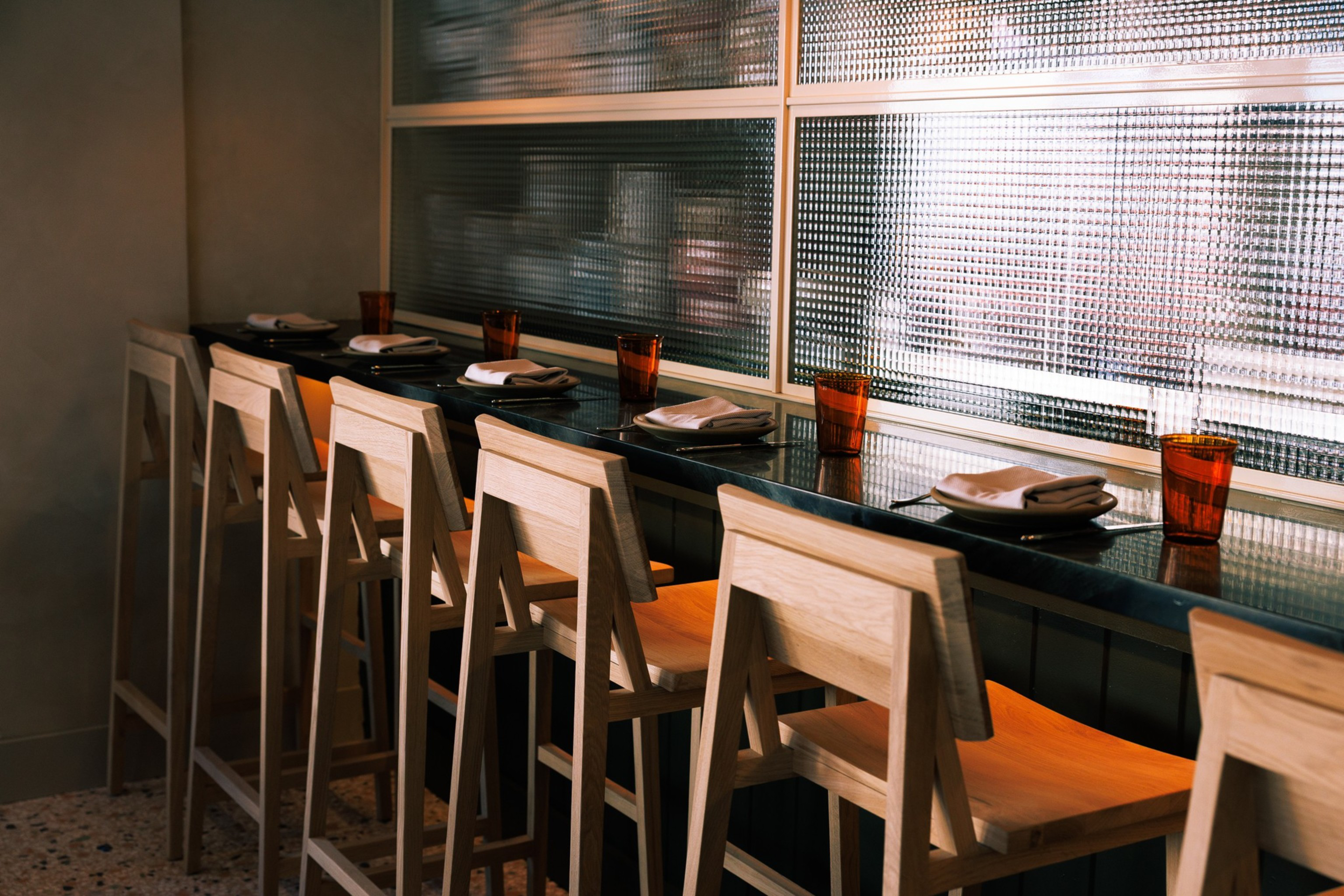 Seven wooden bar stools are lined up at a dark counter with orange ...
