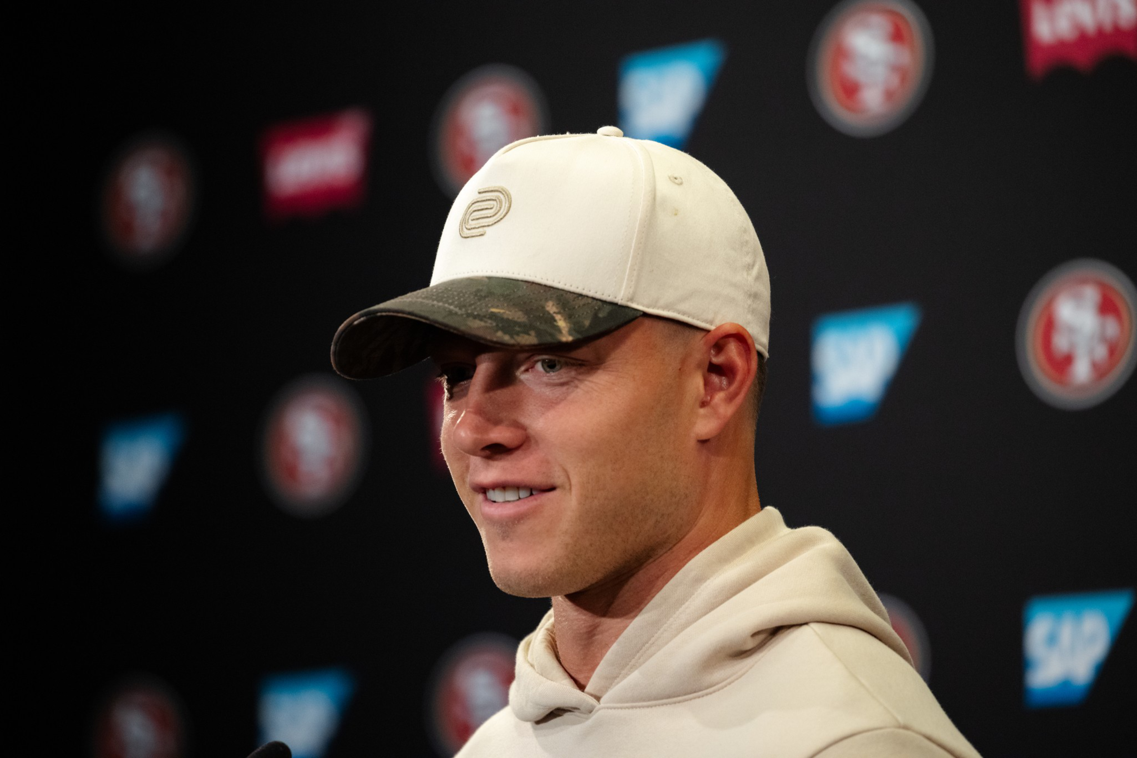 A man in a light hoodie and a white and camouflage baseball cap smiles ...